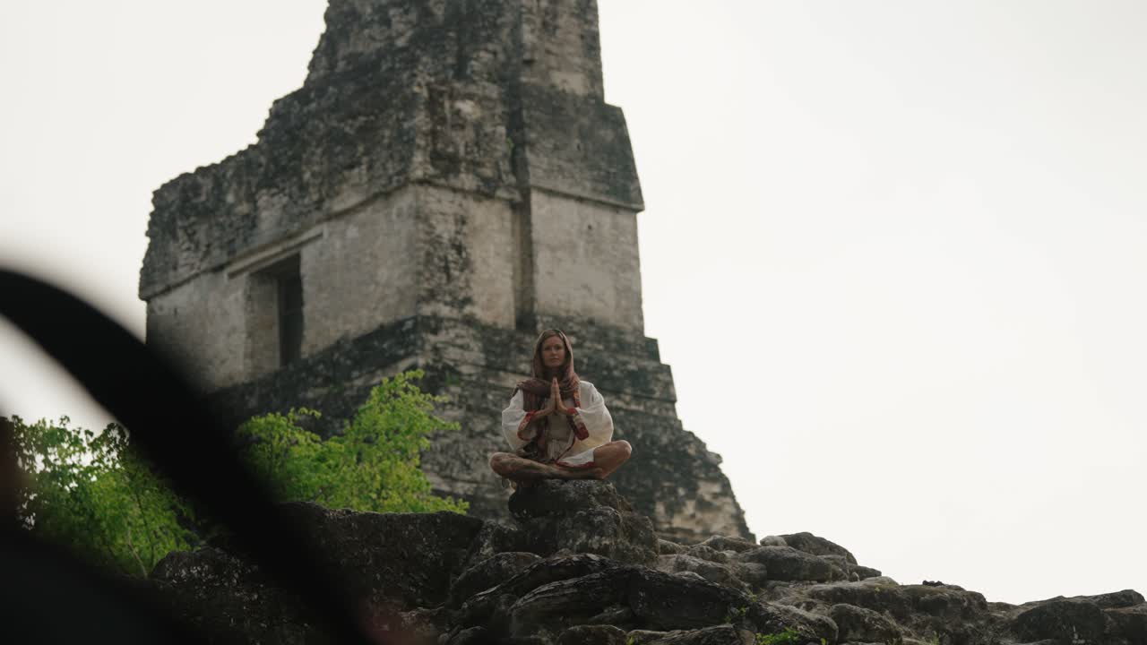 A woman sits cross-legged in meditative prayer pose before an ancient ...