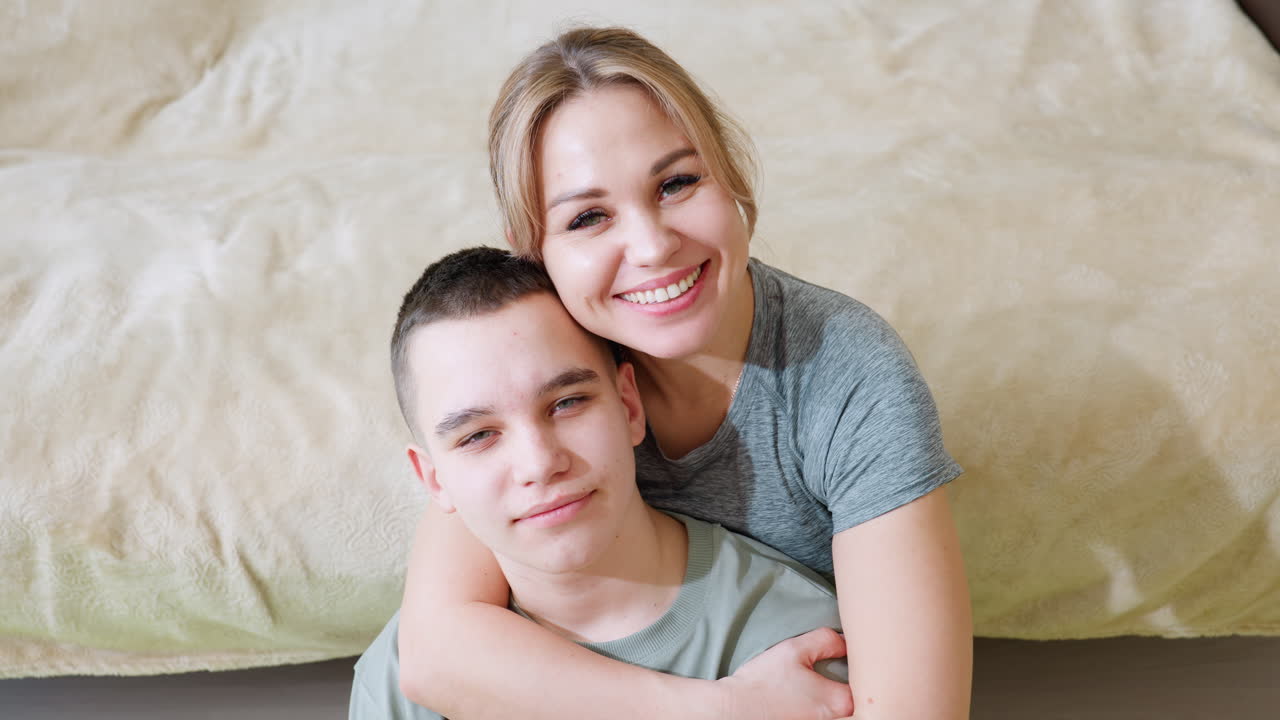 Smiling woman warmly hugs cheerful boy while sitting on bedroom floor ...