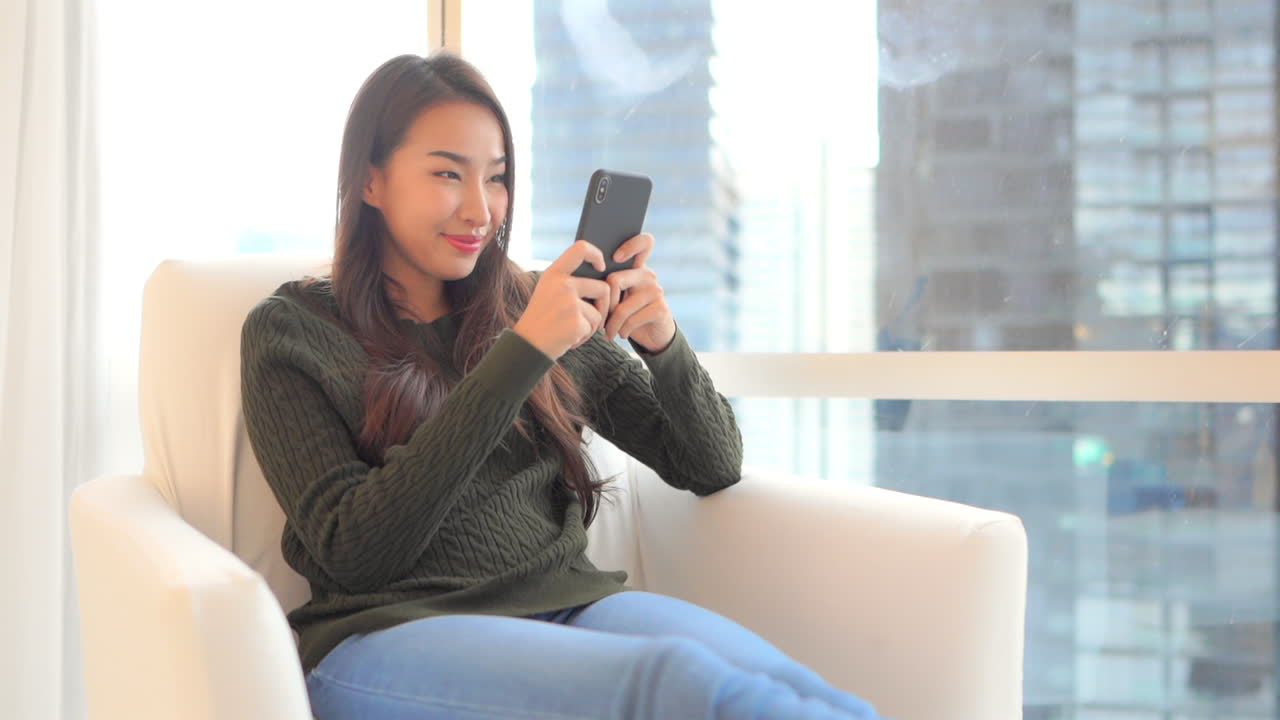 Asian Woman Sitting In Chair By The Window And Texting With Smile On ...