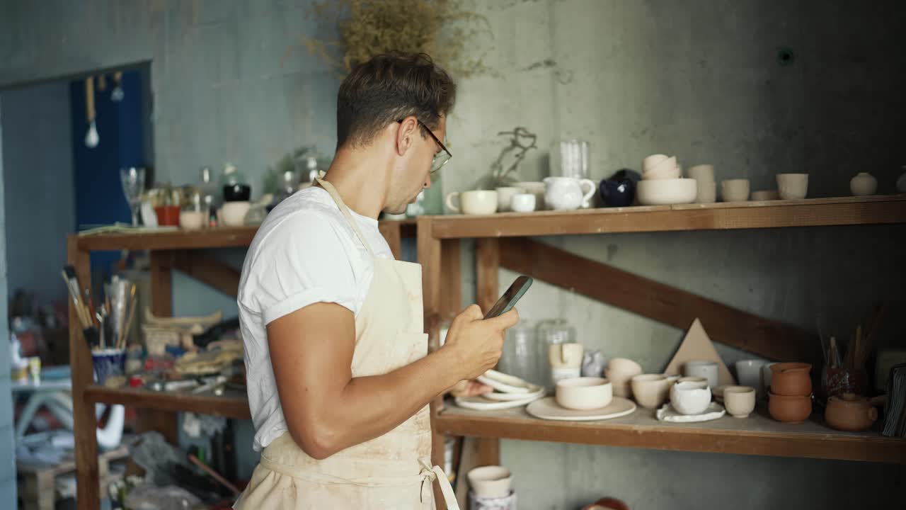 Young happy potter photographing his clay pottery using his phone in ...