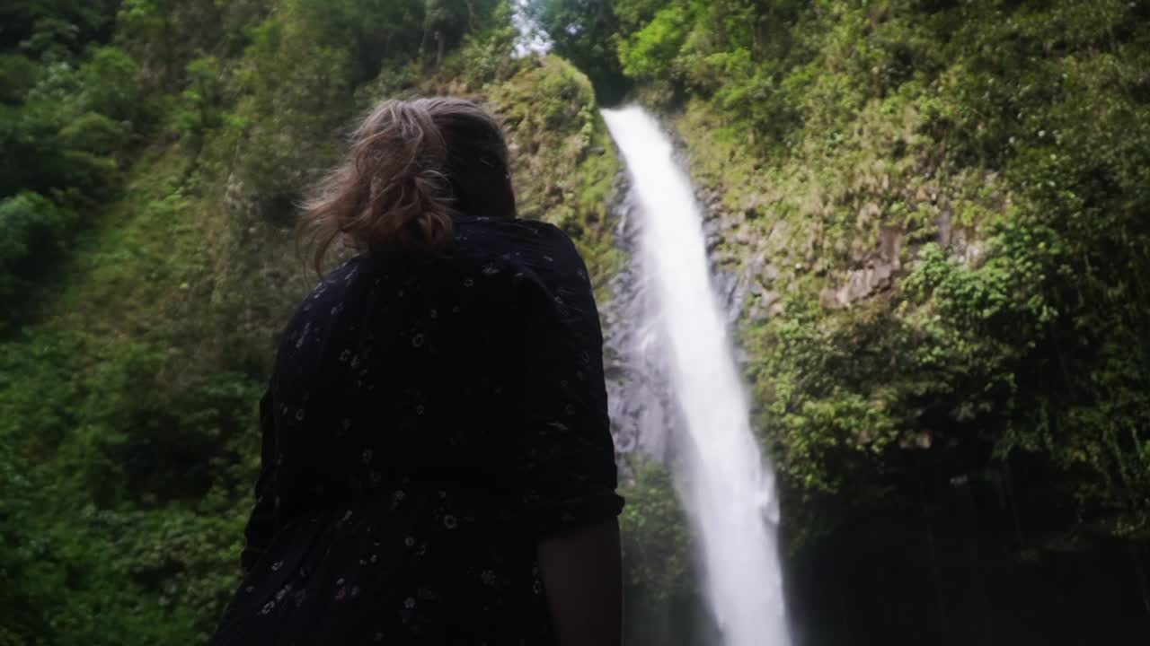 Low angle shot of a women standing in front of a huge waterfall and ...