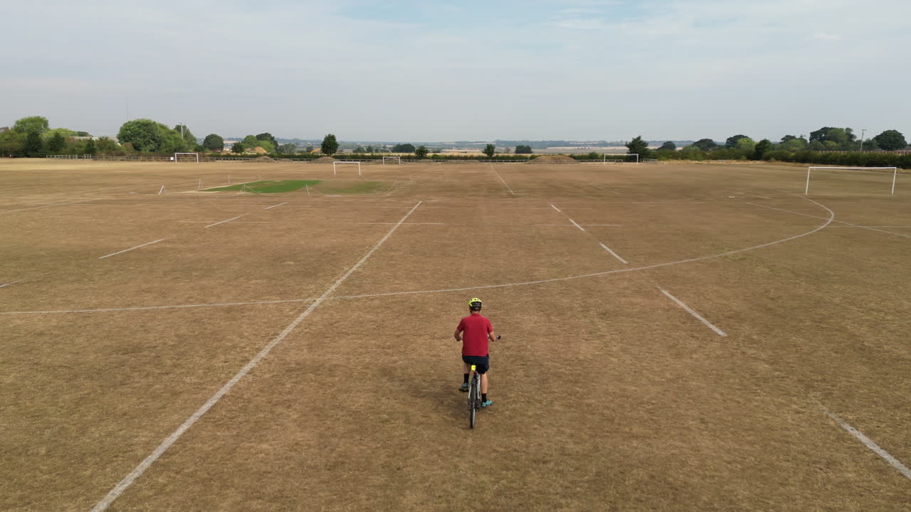 Man With Red Shirt Cycling Across A Playing Field In The UK, Drought ...