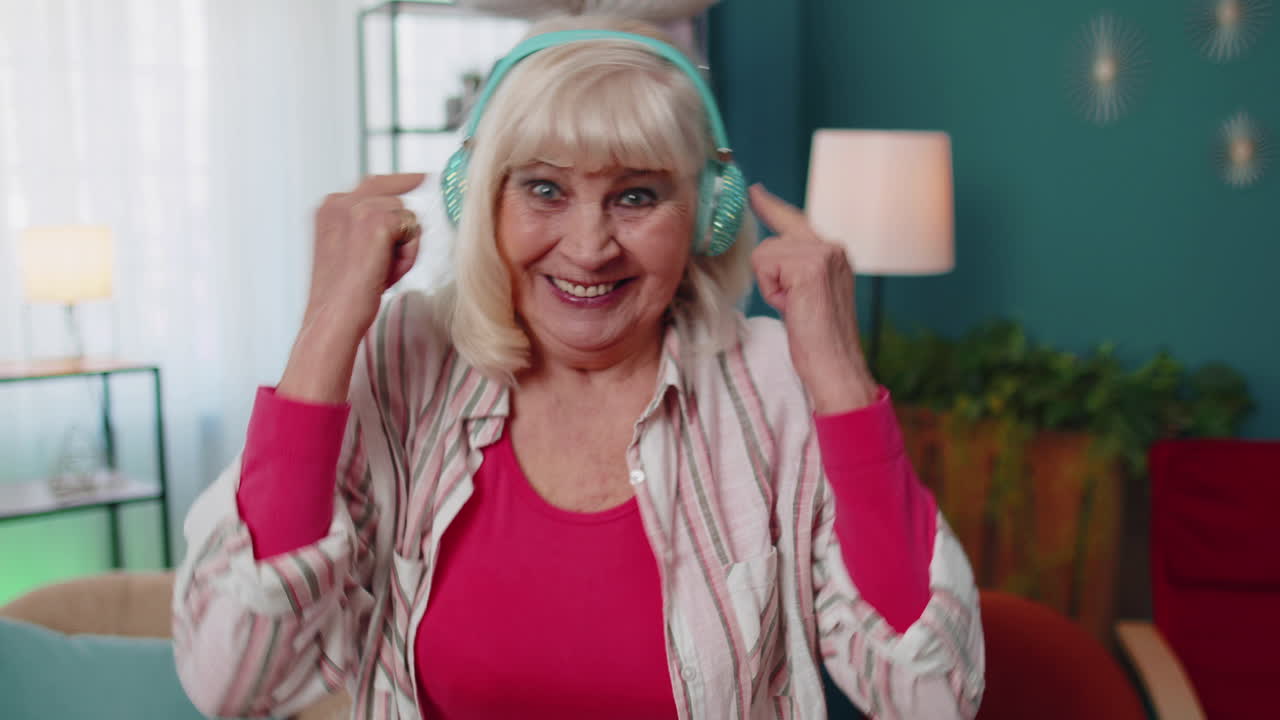 Overjoyed senior woman in wireless headphones dancing, singing on cozy ...