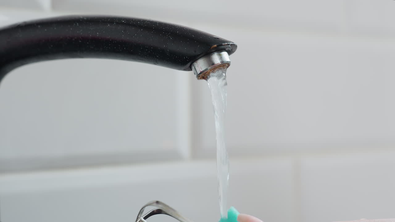 Close-up of running tap with person washing whisk using green sponge ...