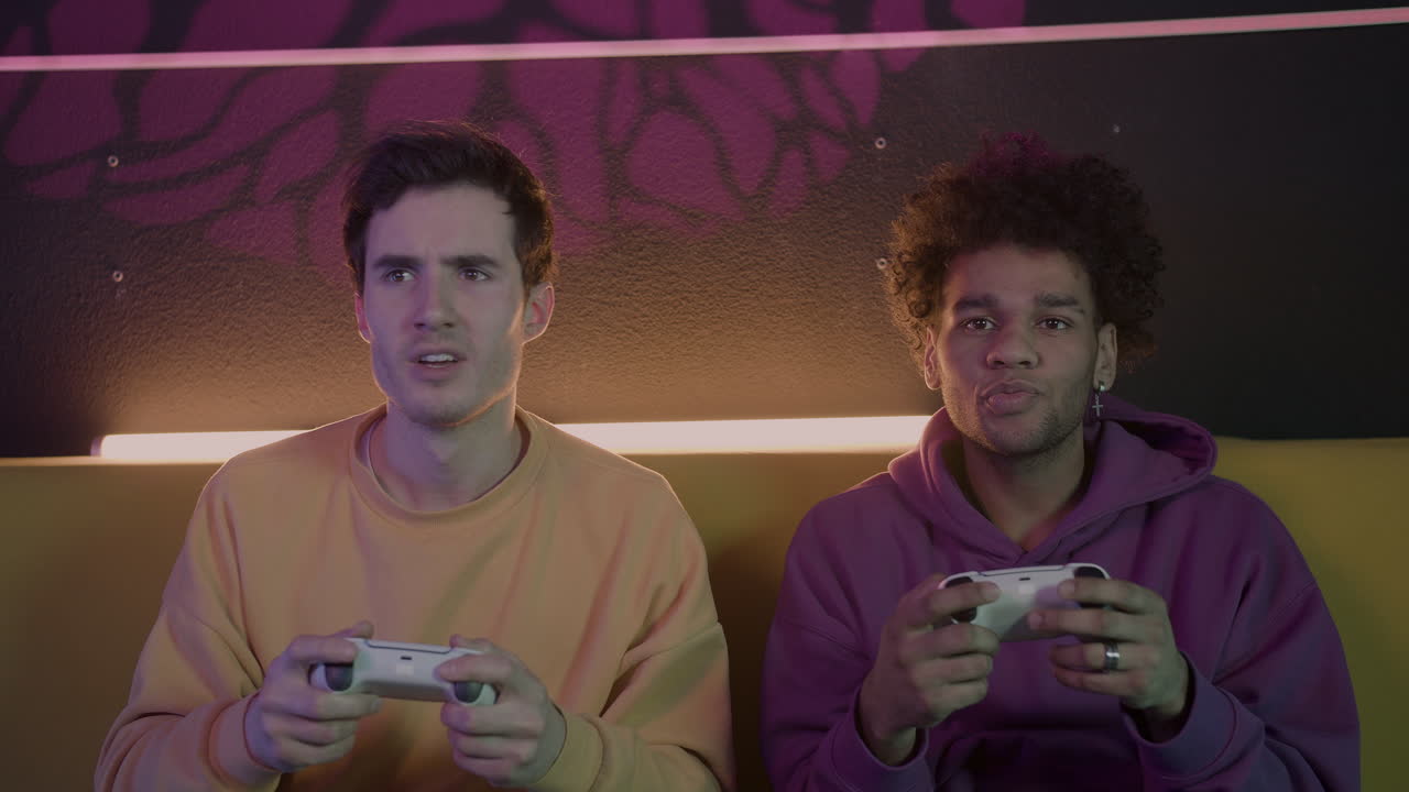 Two concentrated friends playing video games sitting on sofa at home 2 ...