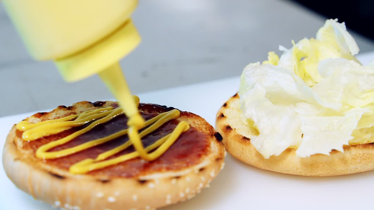 Burger ingredients on table. Pouring mustard on grilled burger bun ...
