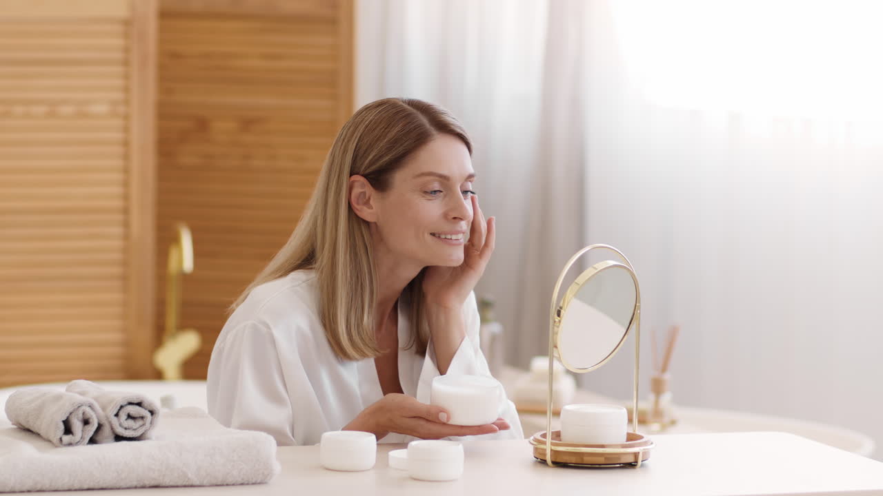 Woman applying face cream in bathroom mirror as part of her skincare ...