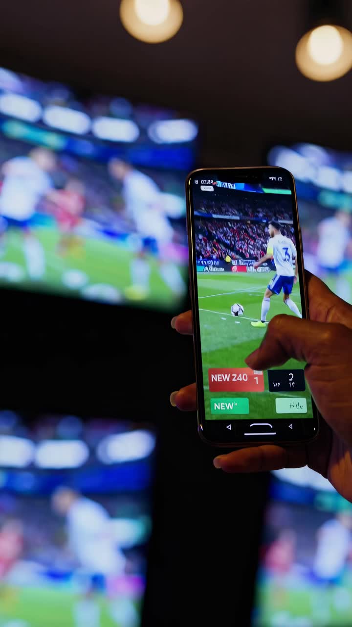 Close-up of a hand holding a smartphone with a sports app, multiple TV ...