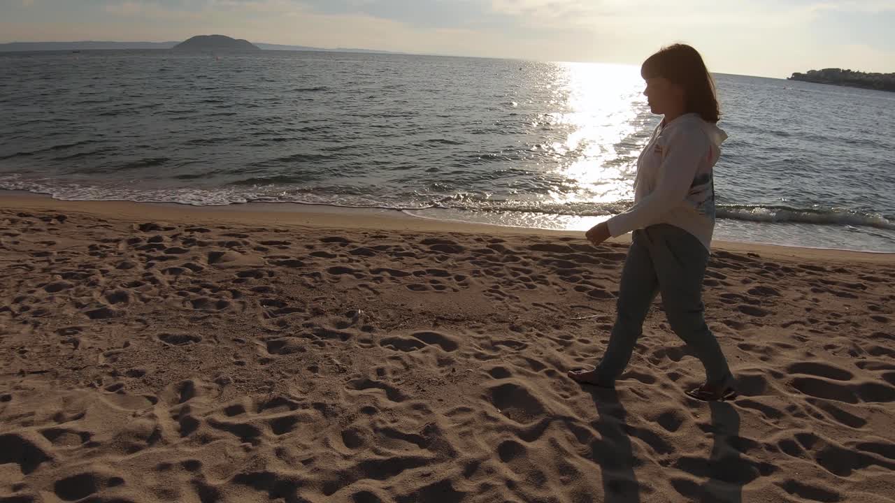 Premium stock video - Girl alone walking on a beach, with sunlight reflection down on a sea ...