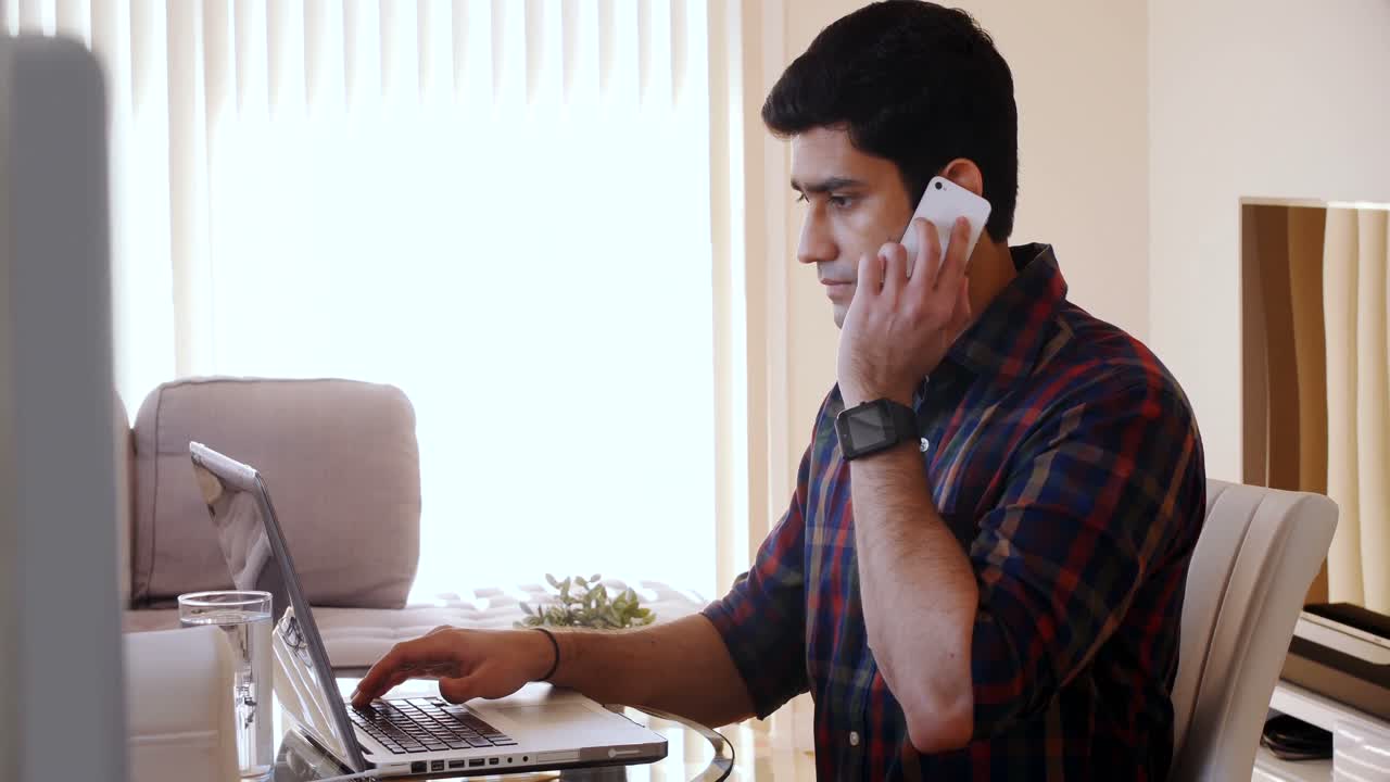 Premium stock video - Man talking on mobile phone while using laptop 4k