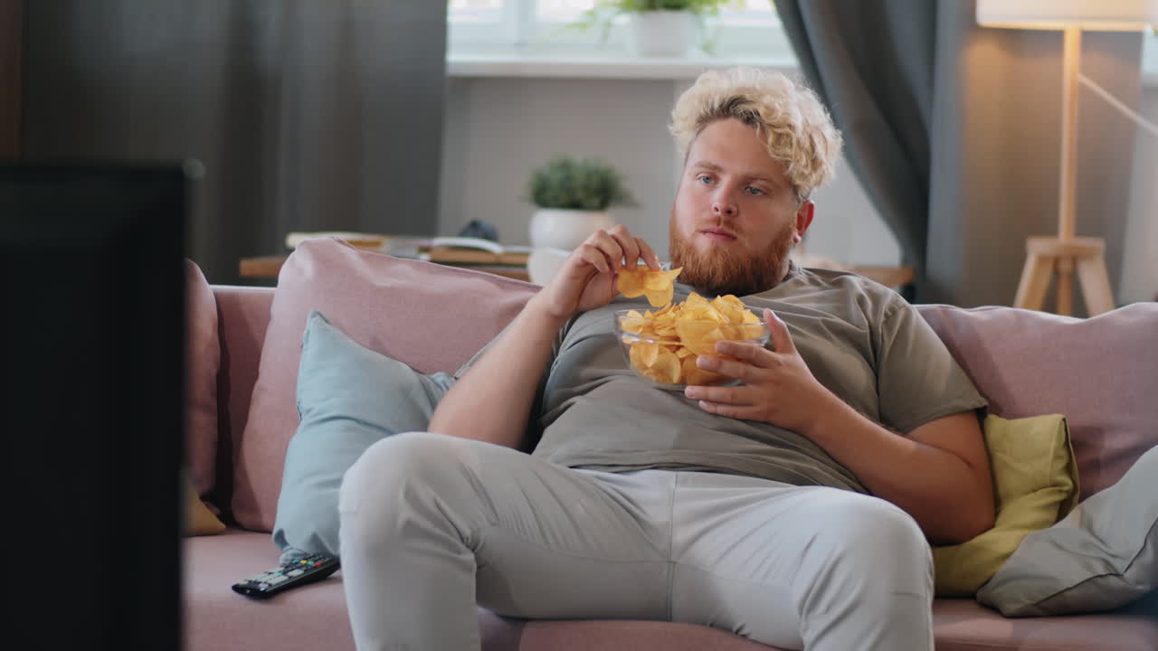 Premium stock video - Obese man eating chips and watching tv at home