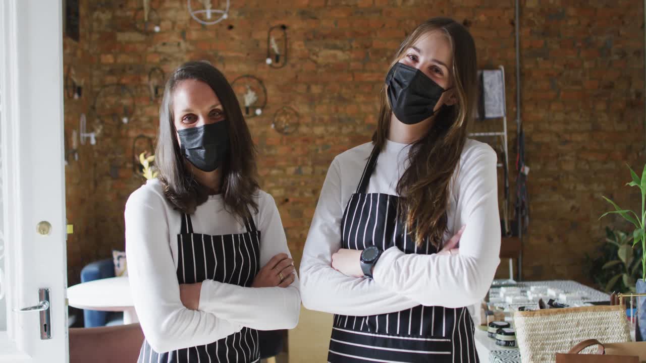 Two caucasian women wearing face masks and aprons, looking at camera ...