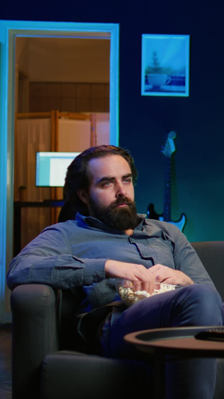 Vertical video sad man on couch eating popcorn from stress while ...