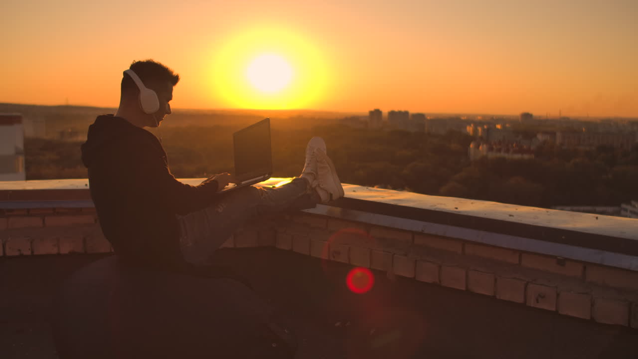 A programmer with a laptop in headphones sits on the roof and writes ...