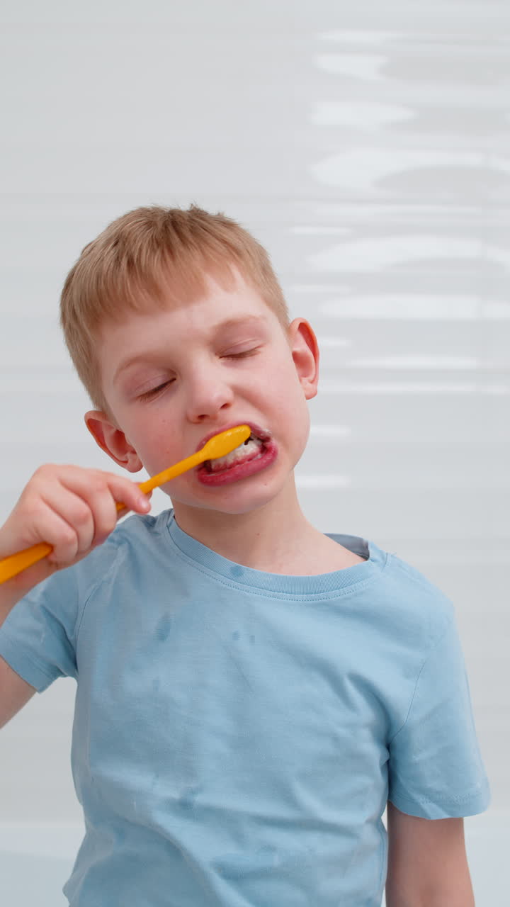 Boy brushing teeth with orange toothbrush smiling slightly in bathroom ...