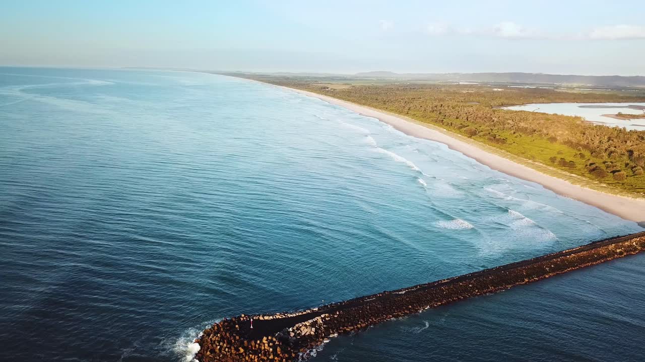 Drone moving side-on showing long straight beach and man made rock wall ...