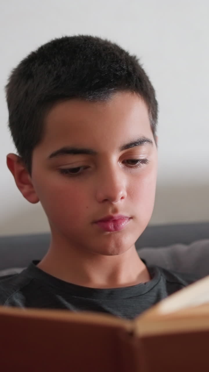 Calm boy deeply focused reading on couch flipping through book pages ...