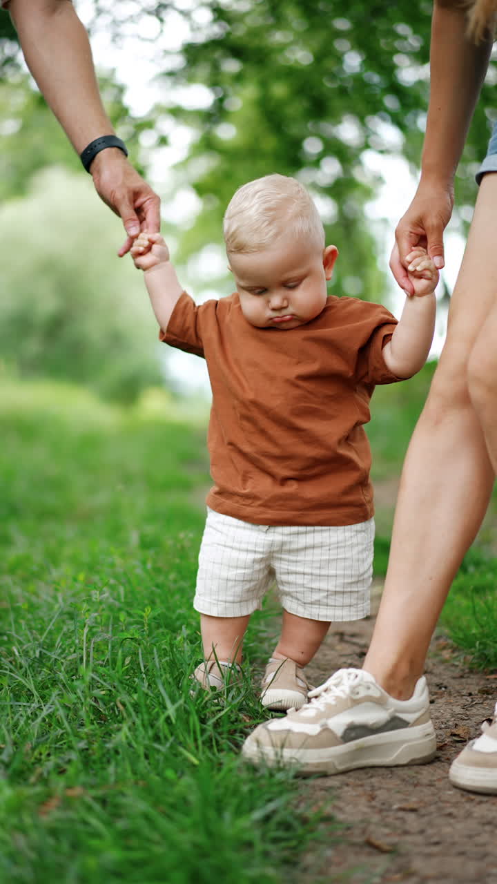 Lovely baby boy holding his parents by the hands looks at their feet ...