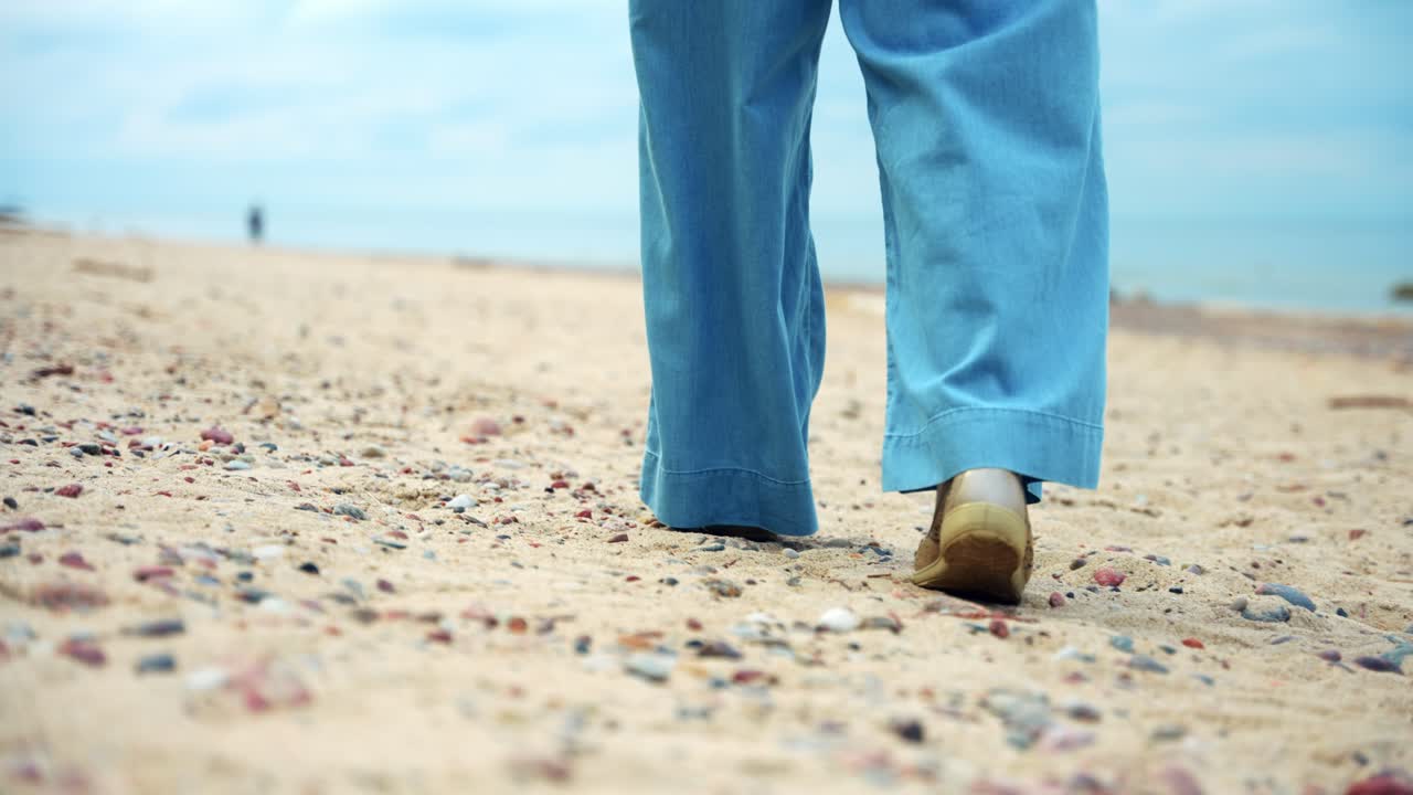 Female wearing blue square pants walks on stony seaside of karkle beach ...